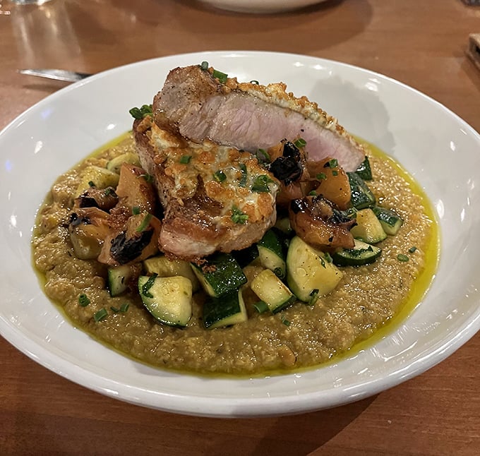 This pork chop resting on a bed of creamy grits with seasonal vegetables is the definition of rustic elegance on a plate.