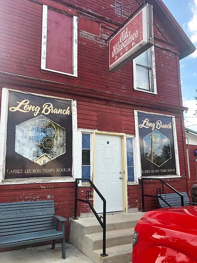 Entrance of a rustic red building labeled "Long Branch," with twin signs featuring a beer logo and the phrase "Laissez Les Bon Temps Rouler," accessed by concrete steps with black railings on either side.