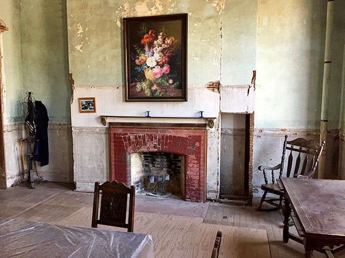 Time stands still in this forgotten parlor where a faded floral painting hangs above a simple brick fireplace—silent witness to decades of history and countless untold stories.