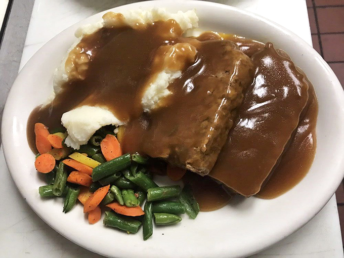 Behold, the legendary meatloaf in its natural habitat – swimming in savory brown gravy, nestled against cloud-like mashed potatoes and colorful vegetables.