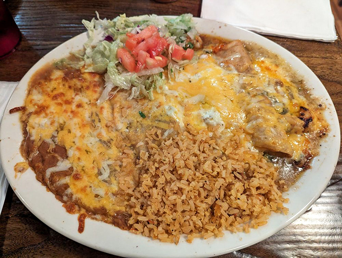 Behold the holy trinity of Mexican comfort: enchiladas, rice, and beans, all unified under a blanket of melted cheese that stretches with each forkful.