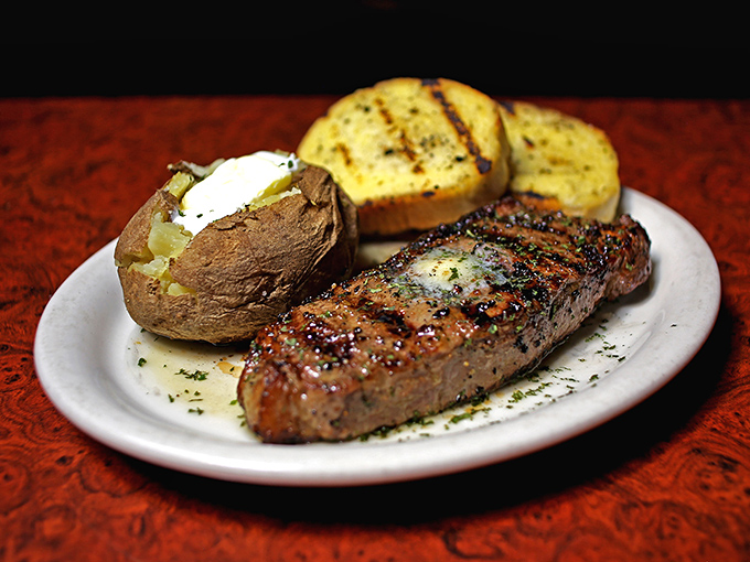 The New York Strip arrives like a work of art, resting beside a butter-laden baked potato and garlic bread—a holy trinity of steakhouse perfection.