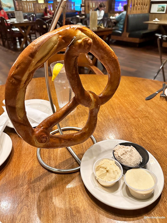 The famous pretzel arrives like edible architecture—hanging majestically on its custom stand with dipping sauces standing by for dunking duty.