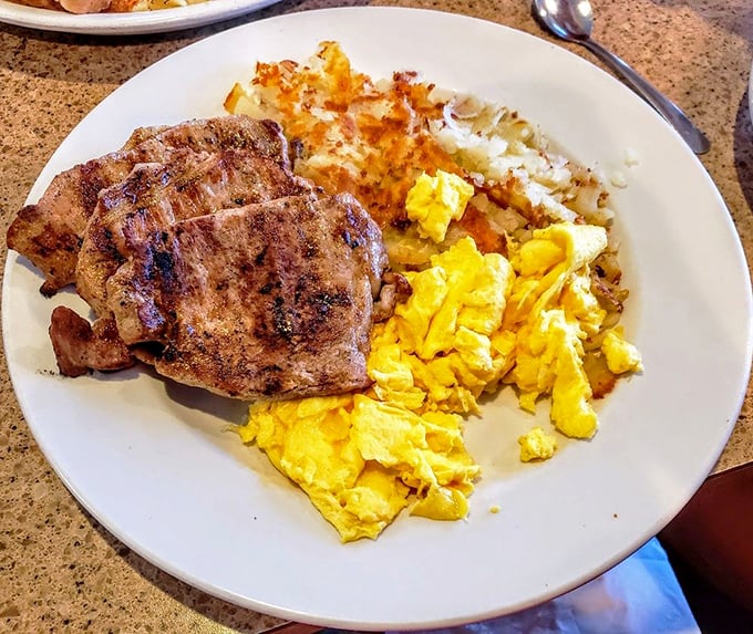 Morning protein done right: savory grilled pork chops sharing plate space with fluffy scrambled eggs and hash browns that achieved the perfect crisp-tender balance.