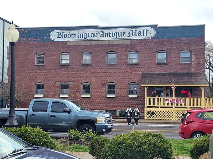 The brick facade of Bloomington Antique Mall stands like a time capsule with its cheerful yellow porch proudly announcing "WE ARE OPEN!" – an invitation to adventure.