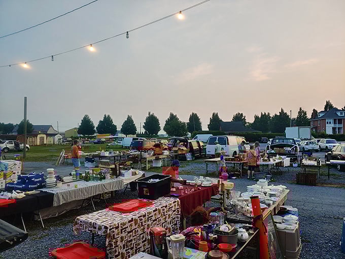 Treasure hunting begins! Tables overflow with a variety of household goods and other items as shoppers browse an outdoor flea market under a dusky sky with string lights overhead. 
