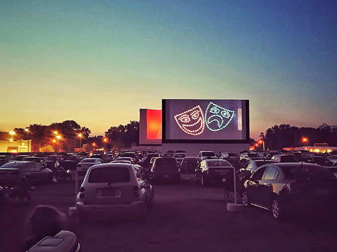 Twilight magic at its finest—cars gathered under a darkening sky as comedy and tragedy masks illuminate the massive screen at Skyview Drive-In.