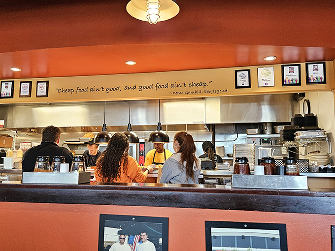 The kitchen pass—where barbecue magic happens beneath a sign that speaks undeniable truth: "Cheap food ain't good, and good food ain't cheap."