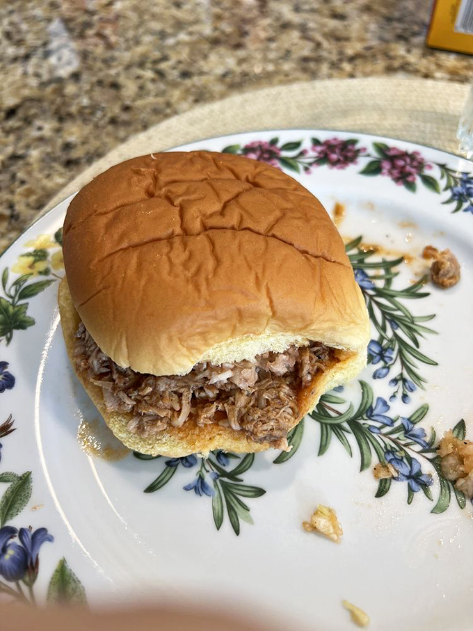 The pulled pork sandwich in its natural habitat—a soft bun on a floral plate, waiting patiently to change someone's definition of perfection.