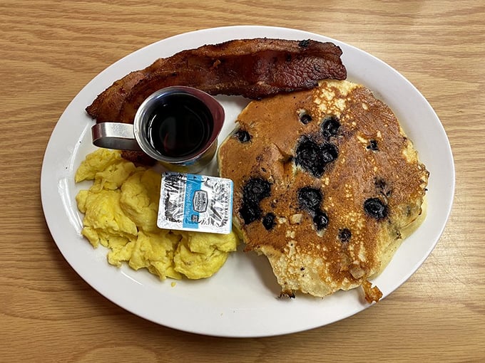 The breakfast trinity: golden blueberry pancake, perfectly scrambled eggs, and bacon that shatters like glass. Heaven on a plate.