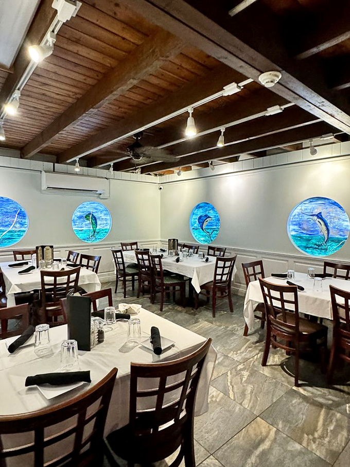 Porthole windows frame marine life paintings like living postcards. White tablecloths and wooden chairs create the perfect backdrop for seafood storytelling.