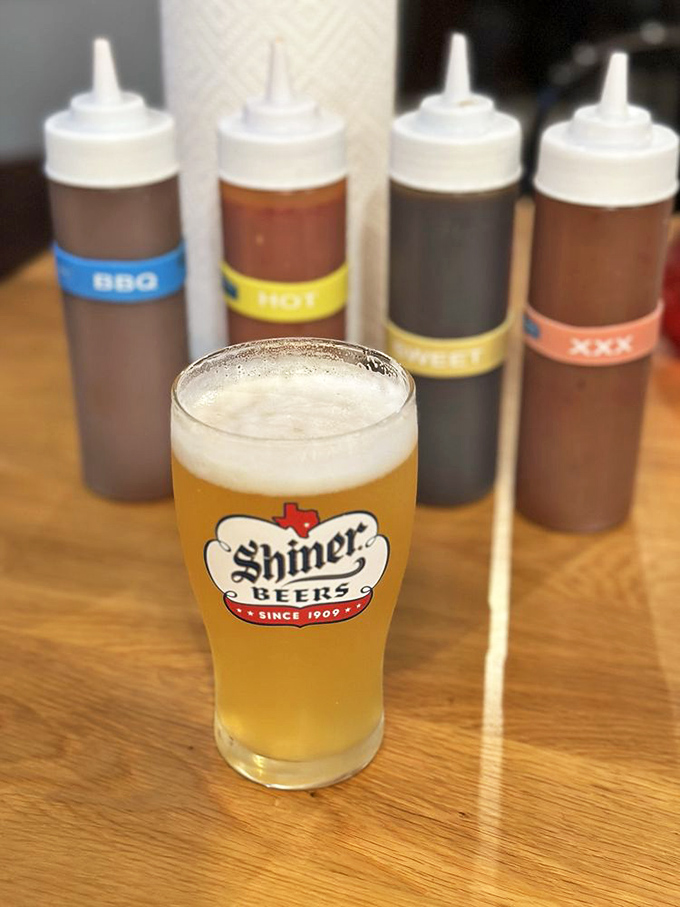 A frosty Shiner Beer standing guard over sauce bottles—because great barbecue deserves a proper liquid companion on its journey.