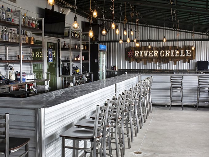 Industrial chic meets comfort at River Grille's bar, where pendant lighting casts a warm glow over metal stools and a sleek countertop.