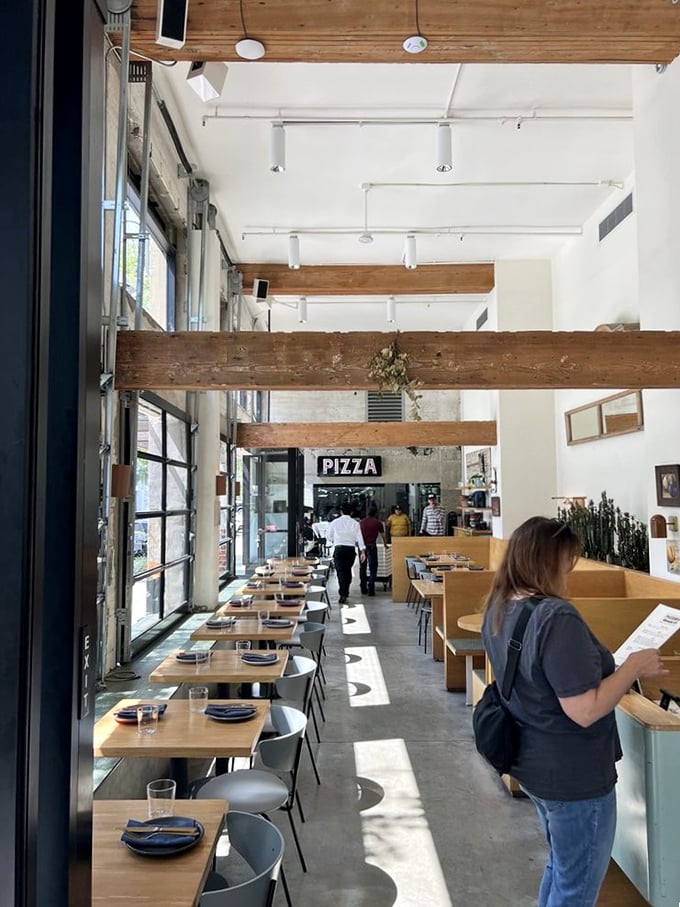 Sunlight streams through floor-to-ceiling windows, illuminating wooden beams and minimalist tables. The restaurant's design philosophy: let the pizza be the star.