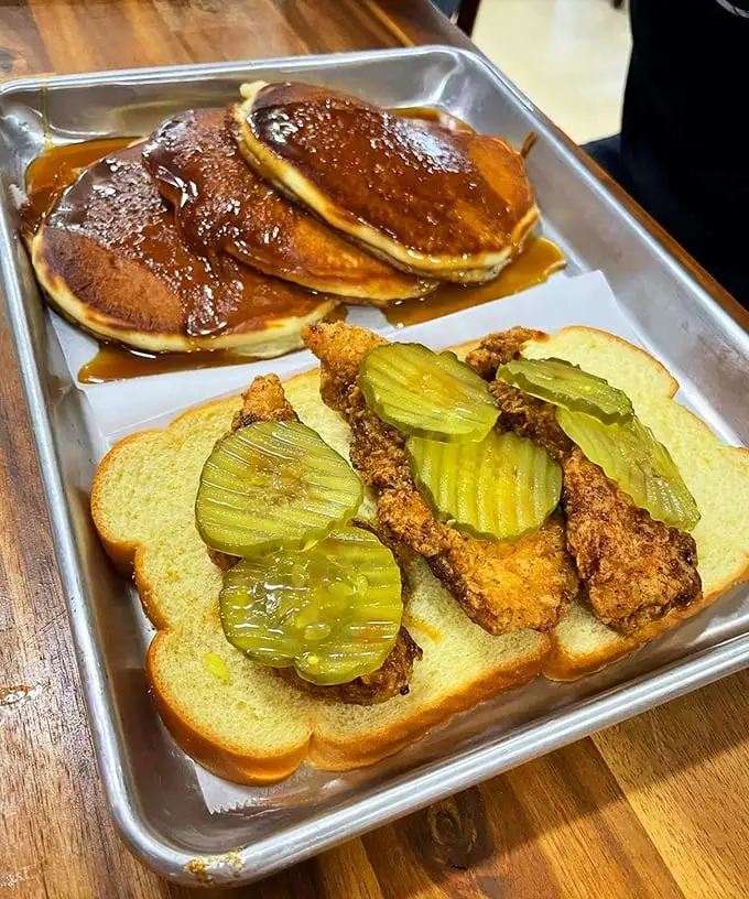 Pancakes drowning in syrup next to fried chicken on white bread with pickles—breakfast and lunch having a delicious identity crisis that works magnificently.