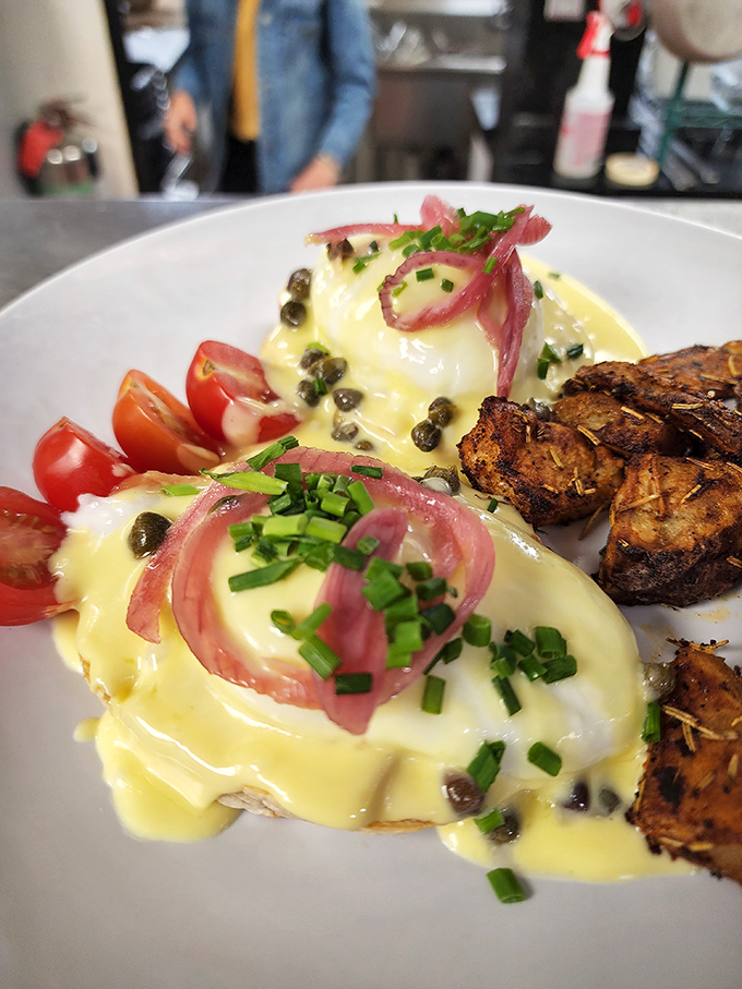 Eggs Benedict reimagined with artistic flair – hollandaise cascading like a golden waterfall over perfectly poached eggs, garnished with capers and pickled onions.