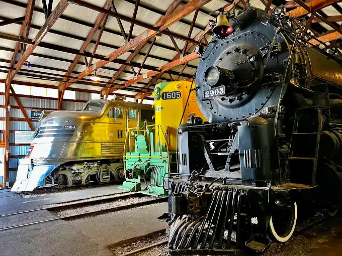 Three legends under one roof: a gleaming silver streamlined engine, a vibrant yellow locomotive, and a powerful black steam engine stand ready, like magnificent time machines waiting to tell tales of railroading's golden eras.