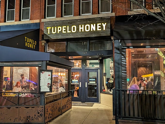 Night falls on Tupelo Honey's warm windows – inside, honey-drizzled chicken works its magic while Asheville's nightlife buzzes outside.