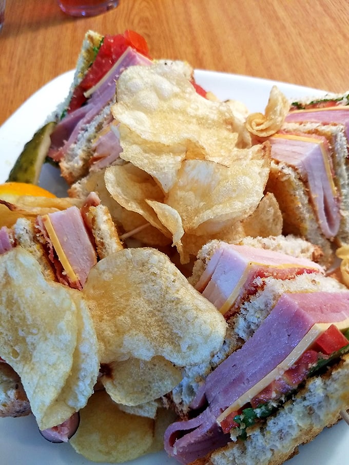 A club sandwich that's architectural brilliance—layers of ham, turkey, and fresh vegetables between homemade bread, with chips standing guard like crispy sentinels.