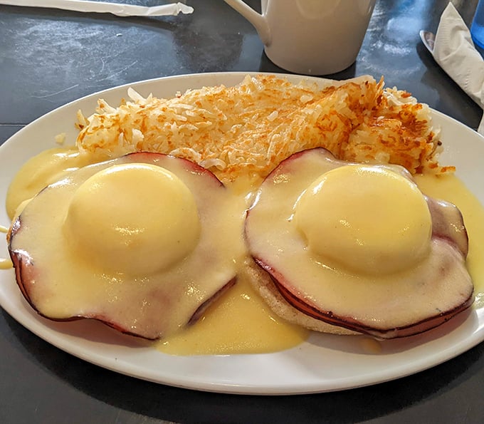 Eggs Benedict reimagined – perfectly poached eggs nestled on Canadian bacon, draped in hollandaise that cascades like a golden waterfall over crispy hash browns.