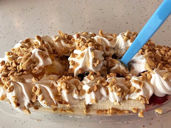 Banana split architecture at its finest—whipped cream mountains, crunchy nut valleys, and that blue spoon standing ready for your excavation expedition.