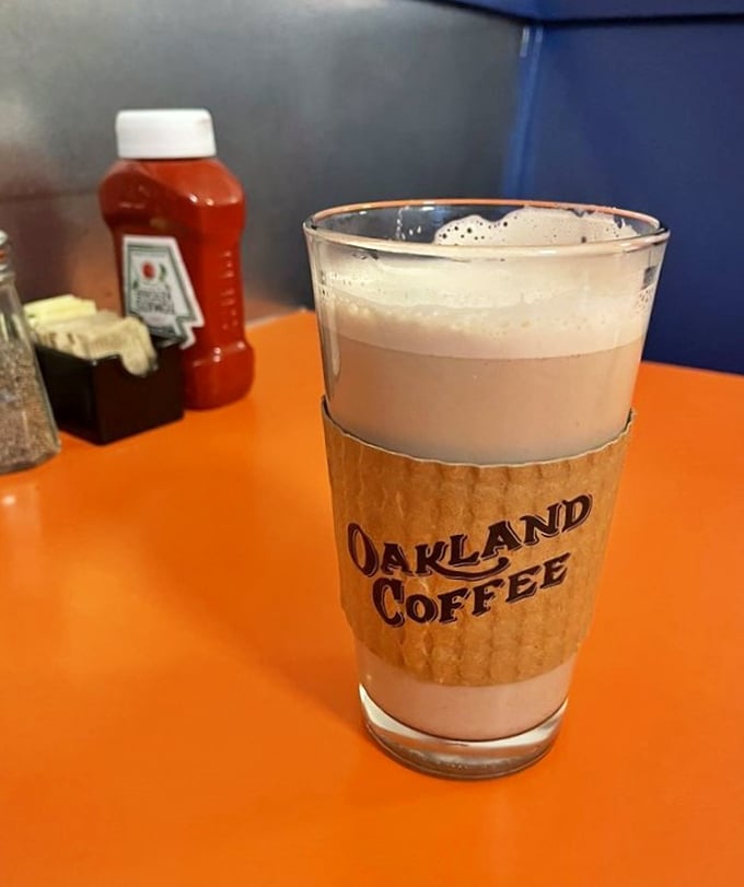 Oakland Coffee in a glass that says "I'm not just caffeine, I'm an experience." The cardboard sleeve isn't just practical—it's a warm handshake.