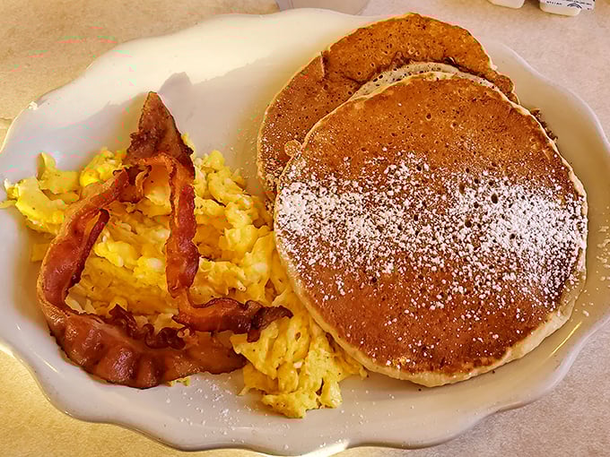 Golden pancakes dusted with powdered sugar alongside scrambled eggs and bacon. This is the breakfast that launched a thousand road trips.