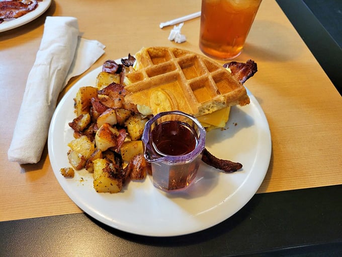Breakfast architecture at its finest—golden waffle creating the perfect foundation for eggs and bacon. The tiny pitcher of syrup adds that touch of breakfast dignity.