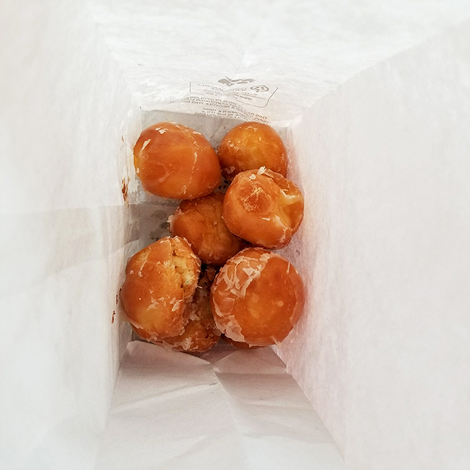 The white paper bag—a humble vessel for pure joy. Six glazed donut holes nestled together like edible pearls of happiness.
