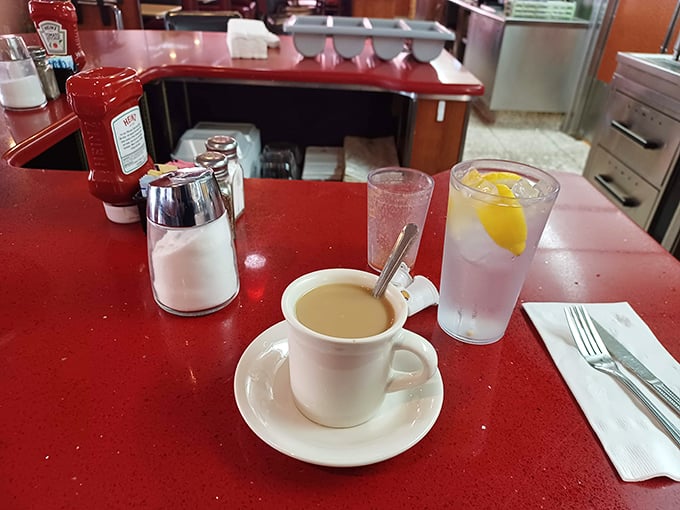 Coffee in a classic white mug on a cherry-red counter—the morning ritual that makes everything possible. Diners understand this sacred truth.