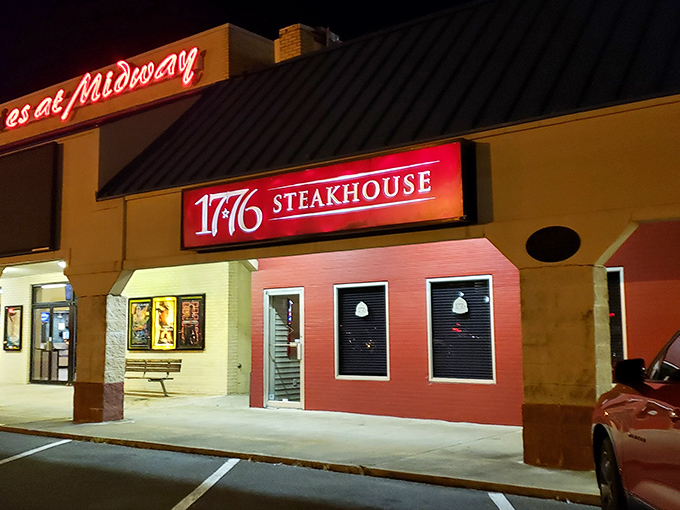 The glowing red sign of 1776 Steakhouse stands out like a beacon for hungry travelers, promising revolutionary flavors in an unassuming shopping center setting.