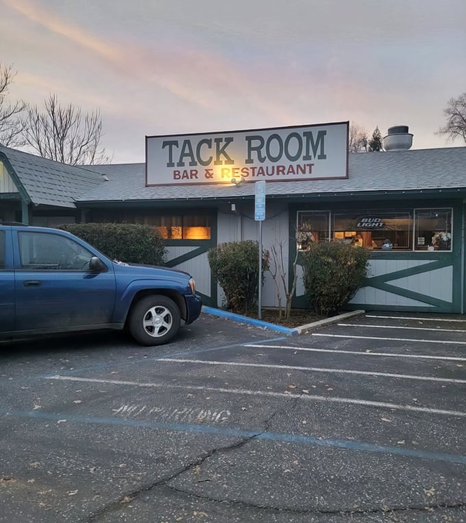The unassuming exterior of Tack Room Bar & Restaurant, where vibrant foliage frames a sign that promises more than it shows—culinary magic awaits inside.