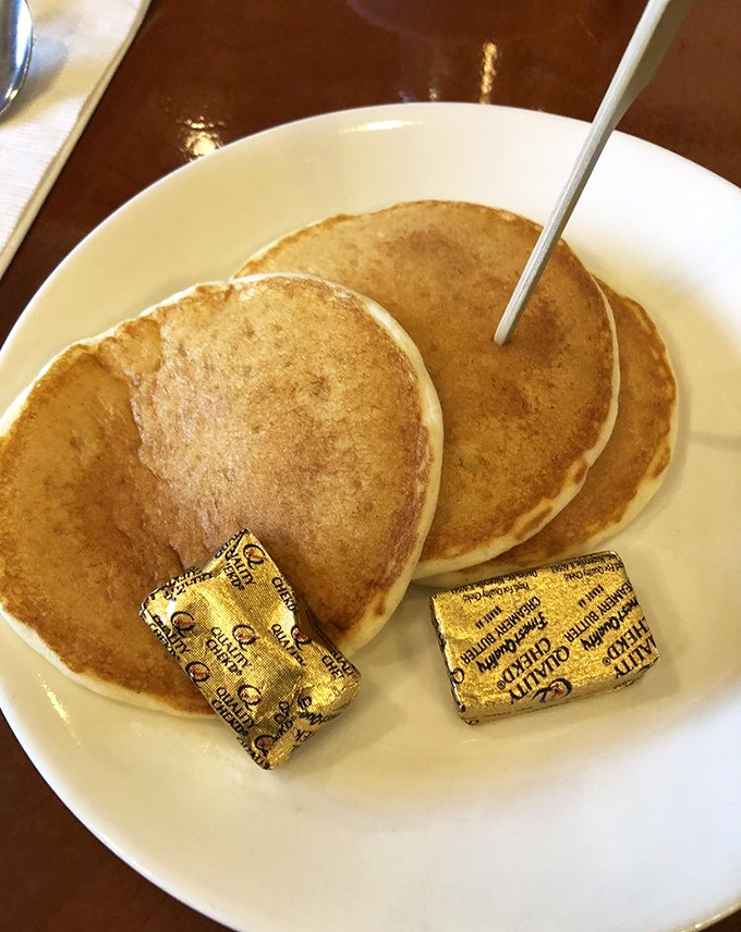 Three perfect pancakes with butter pats still in their gold wrappers—like breakfast presents waiting to be unwrapped and devoured.