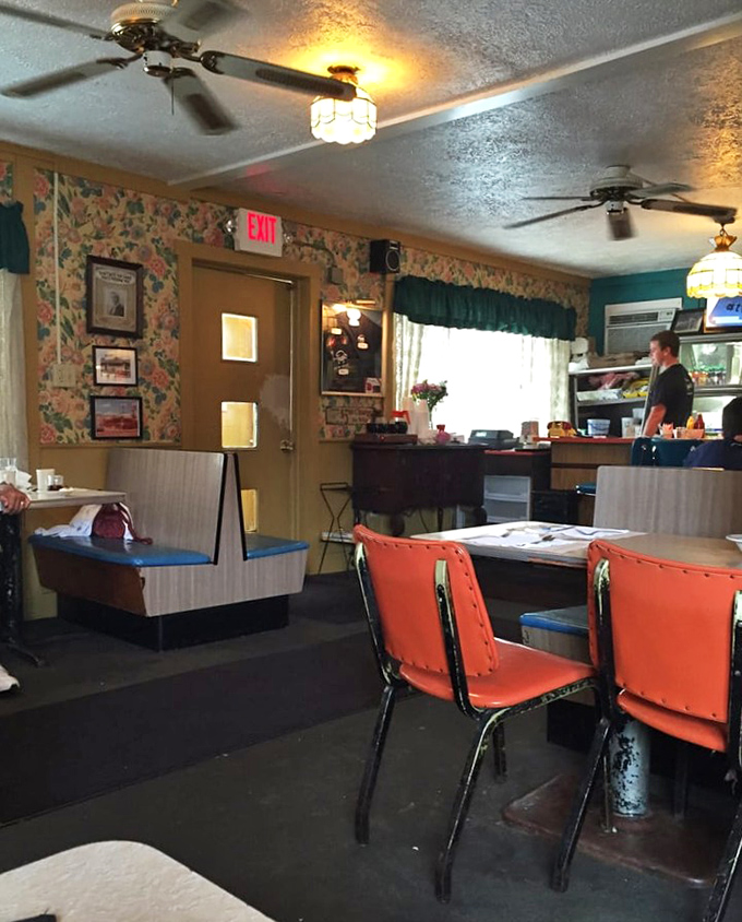 The dining room's charm lies in its unpretentious authenticity – ceiling fans, exit signs, and floral wallpaper creating a time capsule of American dining.