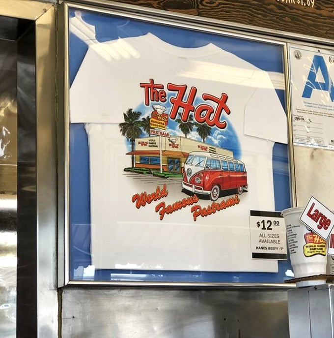 A framed white t-shirt featuring "The Hat" restaurant logo with vintage California imagery including palm trees and a classic VW van, displayed for sale at $12.00.