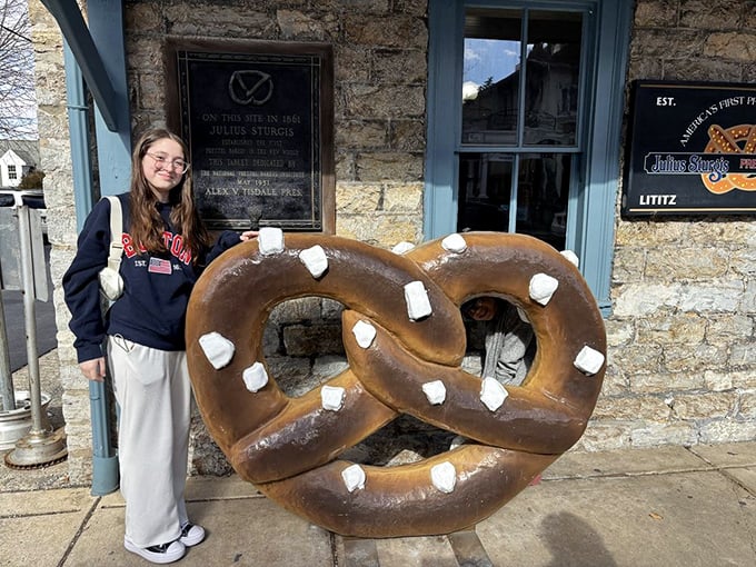 Outside the bakery, a person poses with the giant pretzel sculpture—creating the ultimate carb-lover's holiday card that relatives will actually want to receive.
