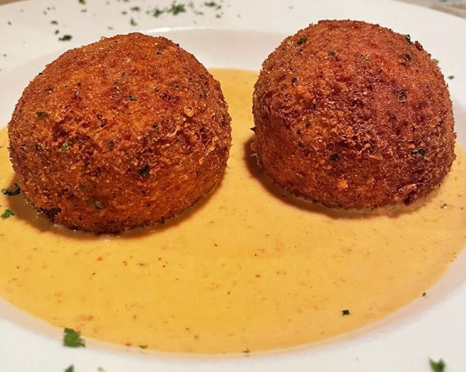 Arancini sitting in a pool of sauce – crispy, golden orbs hiding creamy risotto centers like delicious little worlds waiting to be discovered.