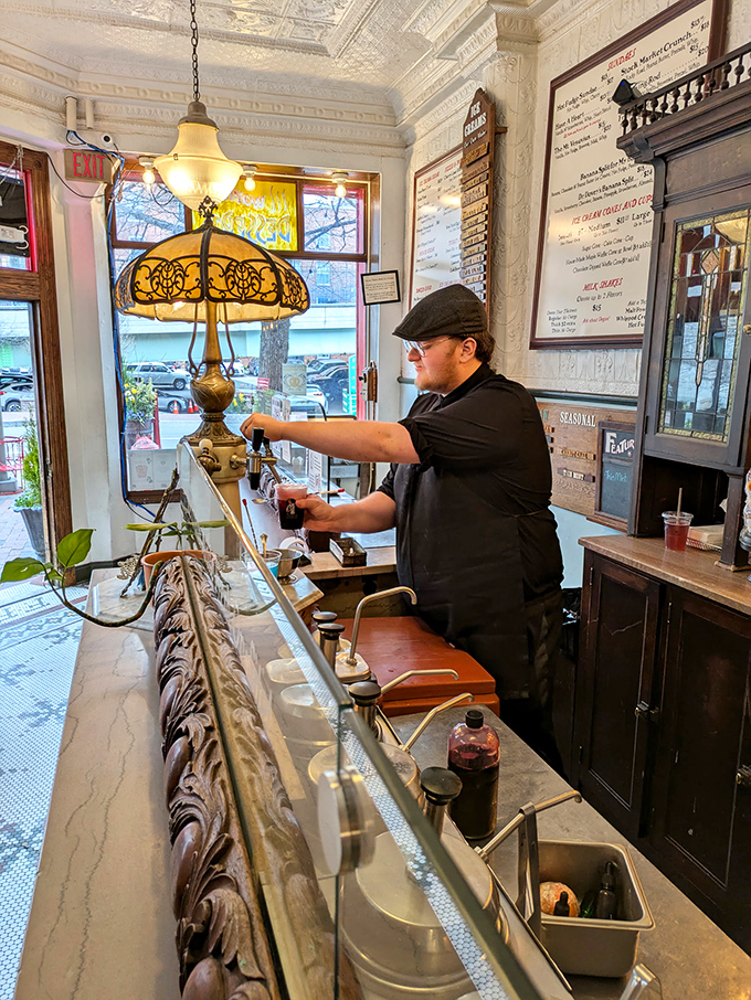 The soda jerk isn't playing dress-up—he's practicing a nearly forgotten American art form. That cap and focused expression speak of craftsmanship, not costume.