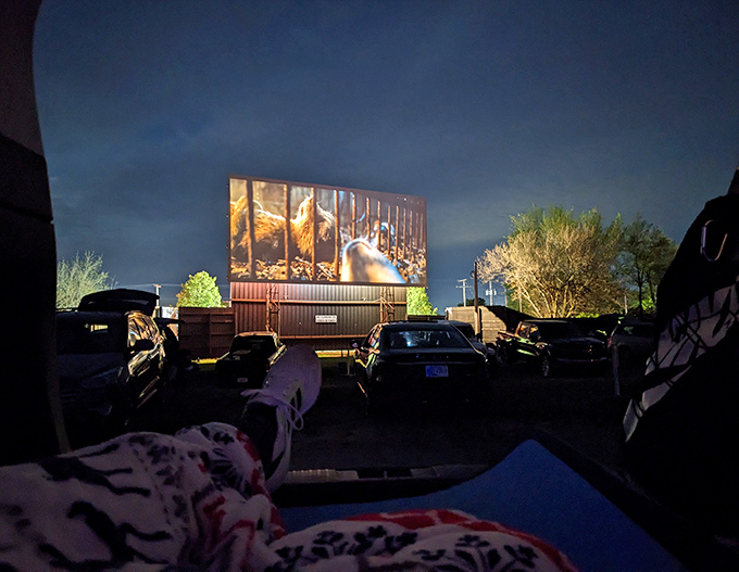 The view from inside a cozy car cocoon – blankets, pillows, and the movie playing across the massive screen. Drive-in perfection captured.