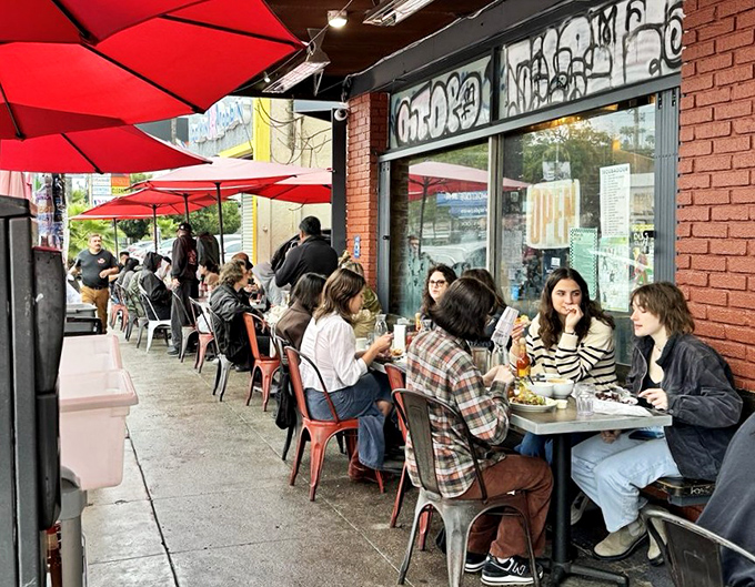 The sidewalk seating scene: where Los Angeles shows up hungry and leaves happy. Red umbrellas create islands of shade for serious breakfast business.