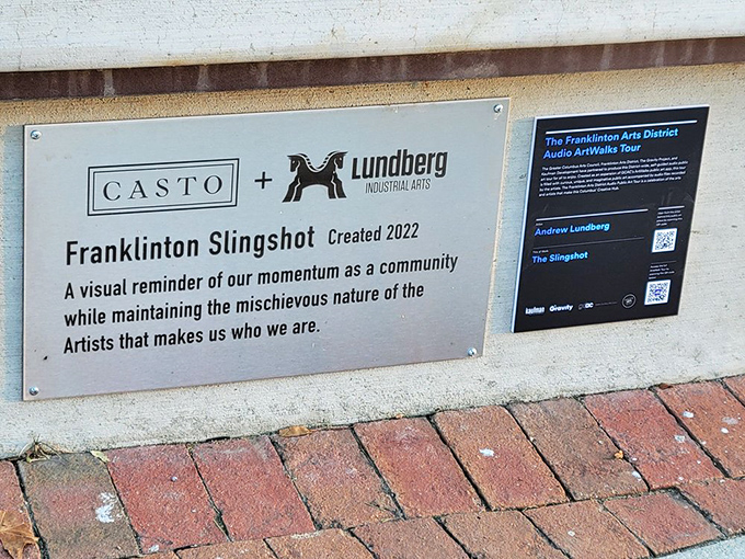 The plaque reveals The Franklinton Slingshot's creation story – a visual reminder of community momentum with a mischievous artistic spirit.
