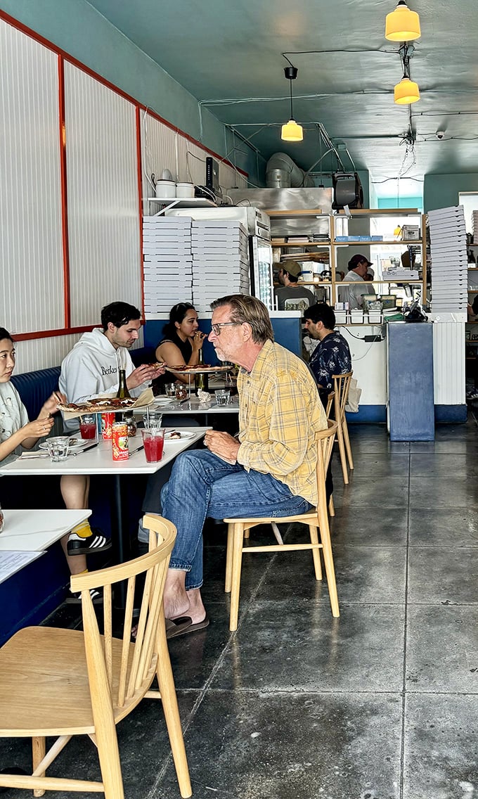 Inside, simplicity reigns supreme with wooden chairs, blue banquettes, and stacks of pizza boxes—a temple where the focus stays squarely on what matters: exceptional pizza.