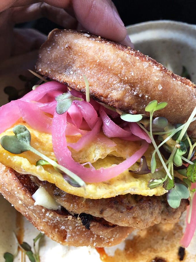 Behold the breakfast sandwich in its natural glory—a perfect storm of fluffy eggs, vibrant pickled onions, and microgreens that make health food feel like rebellion.
