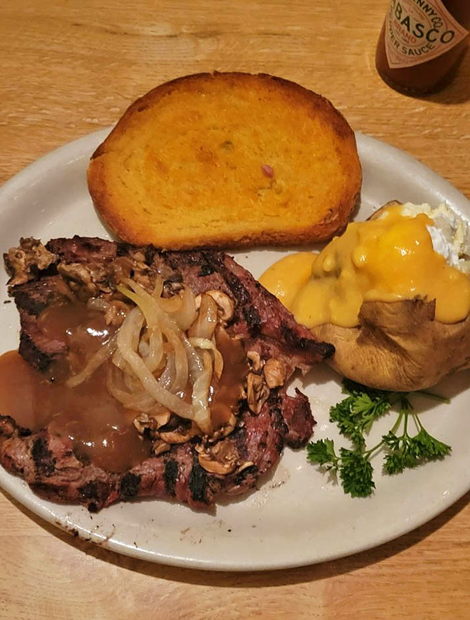Behold the star of the show: a perfectly charred ribeye with sautéed onions, alongside a cheese-smothered baked potato and golden garlic toast.
