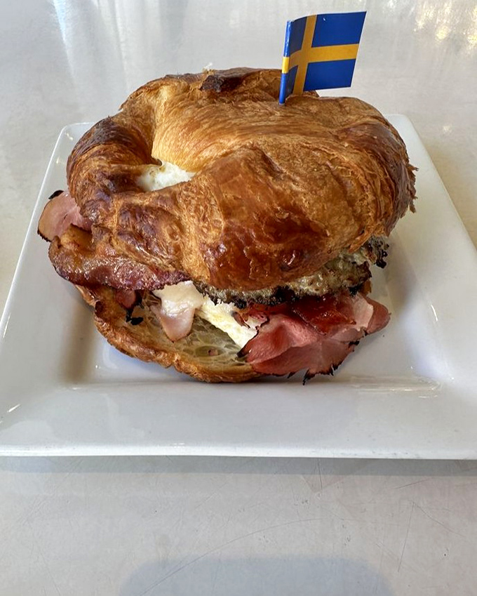 The Swedish flag stands proudly atop this breakfast sandwich masterpiece. It's a morning alliance of flaky croissant and savory fillings that deserves diplomatic recognition.