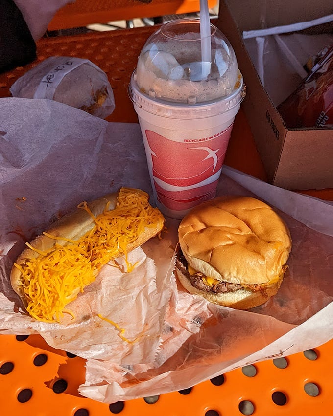 The holy trinity of roadside dining: a juicy cheeseburger, cheese-smothered coney, and a frosty drink—proof that happiness can fit in a cardboard carrier.