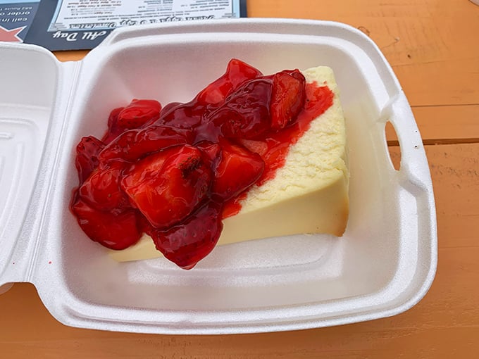 Cheesecake topped with strawberry compote – where dense, creamy decadence meets sweet-tart fruit in a styrofoam container of pure joy.