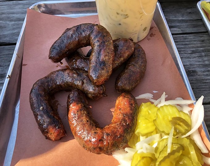 Sausage links curved like smiles, knowing they're about to make your taste buds dance. The pickle garnish isn't decoration—it's necessary balance.