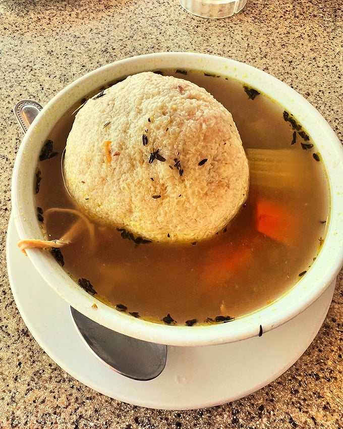 The matzo ball soup—a golden broth cradling what might be the fluffiest cloud of comfort ever to float in a bowl.