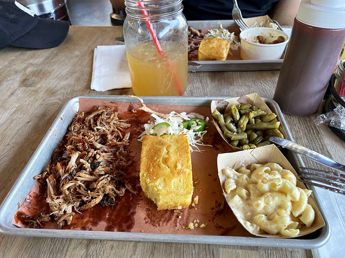 A metal tray loaded with smoky treasures—pulled pork, cornbread, green beans, and mac and cheese. Silverware optional, napkins mandatory.